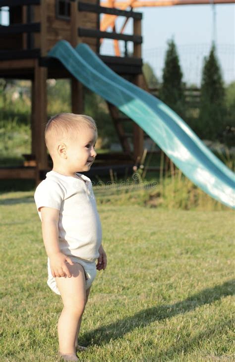 A Small Boy Stands In The Middle Of A Sunny Yard Near A Playground