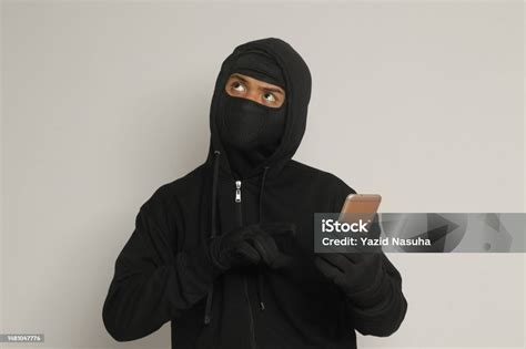 검은 후드티를 입고 휴대폰으로 해킹 활동을 하는 마스크를 쓴 수수께끼의 남자 스마트폰을 들고 있는 해커의 초상화 회색 배경에 격리된 이미지 20 24세에 대한 스톡 사진 및
