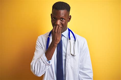 고립 된 노란색 배경 위에 서있는 청진기를 입은 아프리카 계 미국인 의사가 손톱을 물고 손톱을 물고 손으로 스트레스를 받고 긴장합니다 불안 문제 감정에 대한 스톡 사진 및