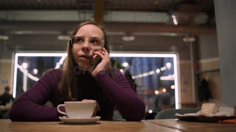 A Woman Is Seated At A Table Engaged In Conversation On Her Cell Phone In The Cafe Stock