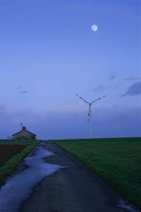 Two Wind Turbines On A Grassy Field At Dusk Near A Rural Road Stock