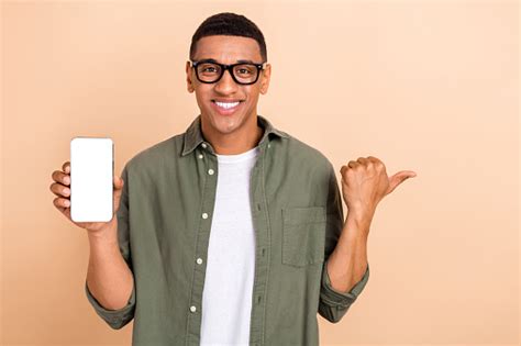 쾌활하고 만족스런 잘 생긴 남자의 초상화는 베이지 색 배경에 고립 된 빈 공간을 지시하는 스마트 폰 터치 스크린을 보여줍니다 Brand Name Smart Phone에 대한