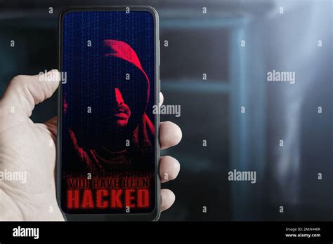 Male Hand Holding Mobile Phone With Hacker Alert And Hooded Cyber Criminal On The Screen
