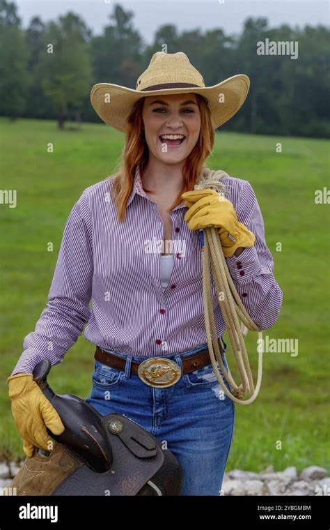 A Beautiful Redhead Model Poses In A Country Setting Wearing Cowgirl Attire And Holding A Saddle