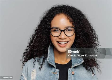 곱슬 머리를 가진 기쁜 여자 광학 안경을 착용 데님 재킷 회색 배경에 고립 된 카메라에 똑바로 보이는 완성 된 작업에 만족