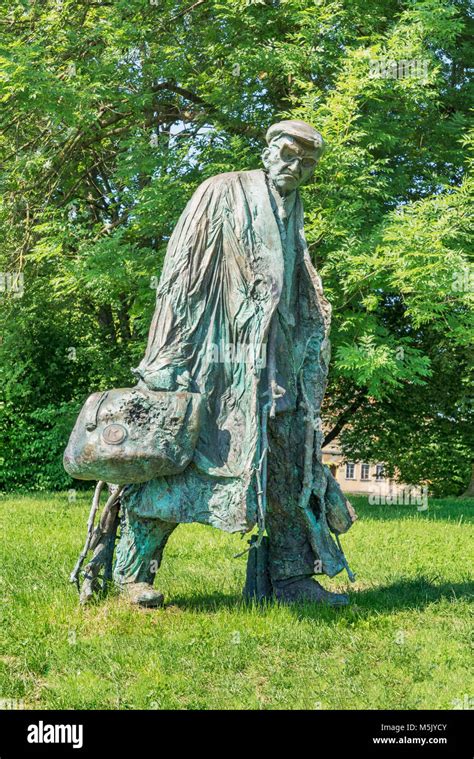 The Bronze Statue Of A Tall Man With A Bag Glasses And Cap Is Located