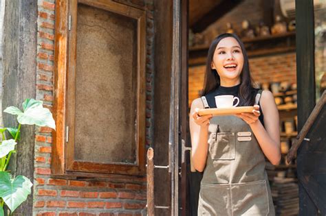 긍정적 인 행복으로 미소 짓는 카페 여성 주인 앞치마 손 선물 트레이와 커피 컵을 가진 아시아 여성 여성이 커피 숍 입구에 서있는 따뜻한 환영 Condident 커피 마실