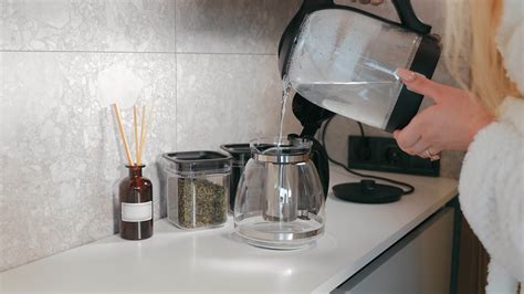 Close Up Of A Person Pouring Water Into A Glass Kettle In A Modern Kitchen Setting Hand Pouring