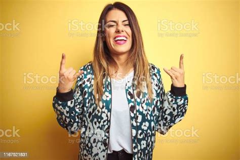 노란색 고립 된 배경 위에 캐주얼 재킷을 입고 젊은 아름다운 여성은 손을 위로 바위 기호를하고 미친 표정으로 외칩니다 음악 스타 무거운 음악 개념 갈색 머리에 대한 스톡