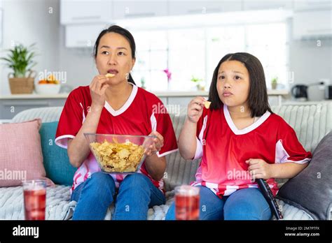 Asian Mother And Daughter In Football Shirts Watching Soccer Game Supporting And Eating Crisps