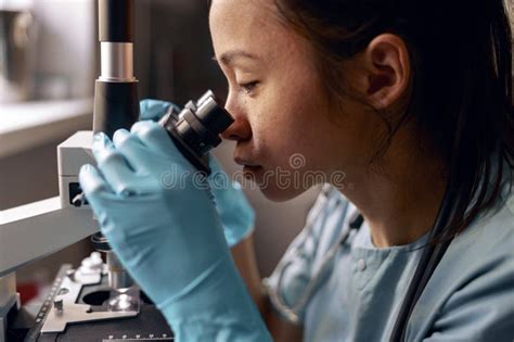 Asian Woman In Blue Gloves Looks Into Microscope Lens Sitting At Workplace In Laboratory Stock