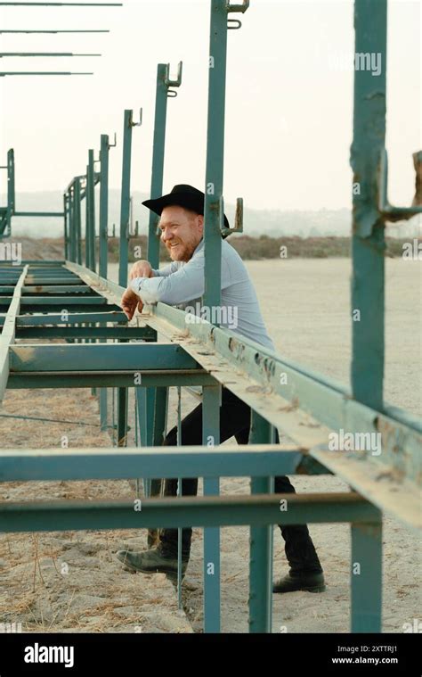 Man In A Cowboy Hat Leaning On A Metal Structure Smiling Outdoors