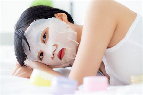 침실에서 침대에 시트 페이셜 마스크를 가진 아름다운 젊은 아시아 여성 미용 소녀는 집에서 주름 한 사람 스킨 케어 치료 및 건강을 위해 피부 관리 얼굴로 화장품과 메이크업을
