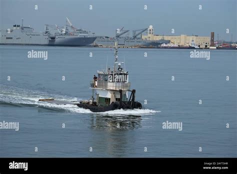 A Small Utility Tug Boat Working On Upper Bay With Navel Ships In The Background Seen From The