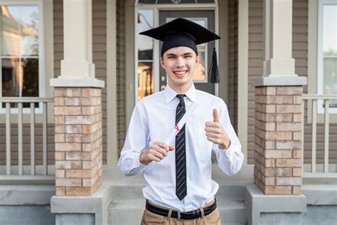 엄지손가락을 들고 집 앞에서 졸업모자를 쓰고 졸업장을 들고 있는 젊은 남성 학생 고등학교에 대한 스톡 사진 및 기타 이미지 고등학교 졸업식 교외 Istock
