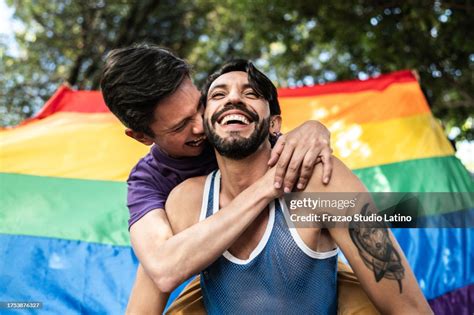 Gay Couple Piggyback Outdoors With Rainbow Flag On Background High Res