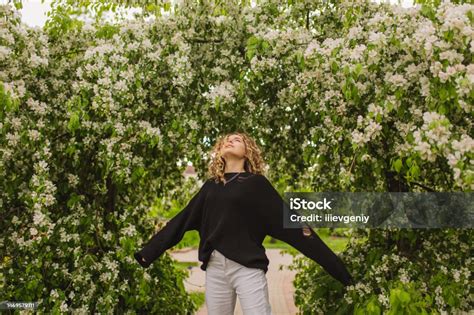 Blonde Curly Girl In Black Sweater On Background Of Flowering Tree