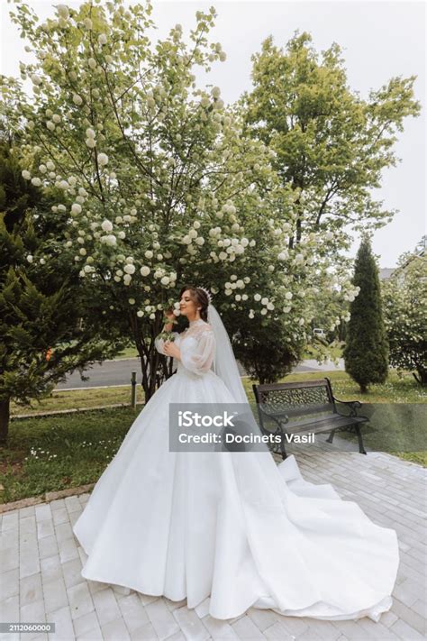 가슴이 트인 레이스 드레스를 입은 곱슬곱슬한 갈색 머리 신부는 무성한 베일을 쓰고 부케를 들고 푸른 나무를 배경으로 포즈를 취합니다 봄 결혼식 20 24세에 대한 스톡 사진