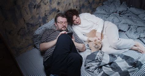 Mature Couple Are Resting In Bed Watching Tv And Stroking The Cat Stock