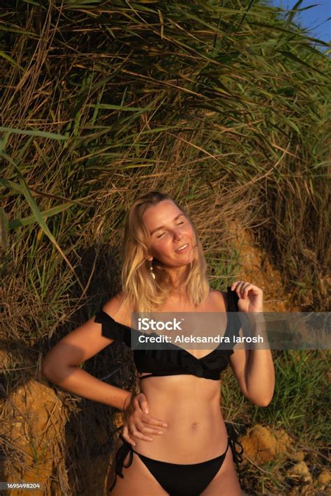 Young Blonde Woman In Black Swimsuit Posing On The Beach At Sunset