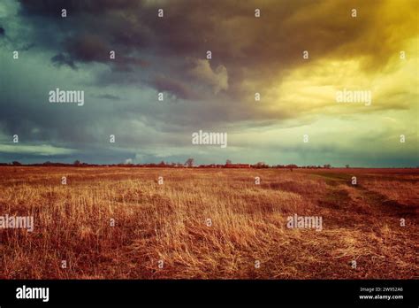 The Image Shows A Vast Field With Dry Grass Under A Dramatic Sky Filled With Dark Clouds