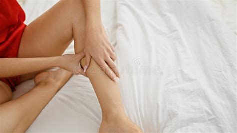 A Young Caucasian Woman In Casual Attire Massages Her Leg On A White Bed In A Home Setting Stock