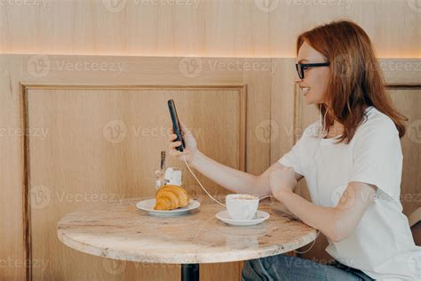 Happy Redhead Young Woman Uses Roaming Connection Sits At Table In
