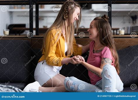 Two Smiling Lesbians Holding Hands While Sitting On Sofa In Living Room