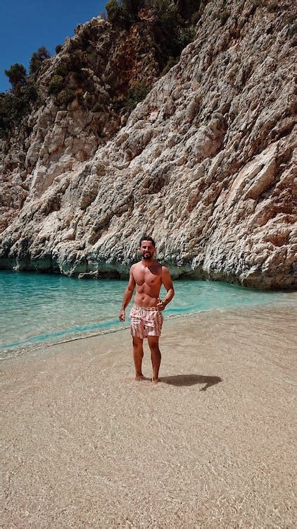 A Topless Man Standing On The Shore Of A Beach Near Big Rock Formation