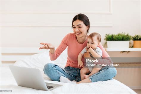 흥분한 어머니는 아기를 안고 노트북으로 화상 통화를 하고 침대에 앉아 컴퓨터 웹 카메라에서 몸짓을 한다 가족에 대한 스톡 사진
