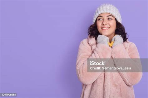 따뜻한 모피 코트와 모자를 쓰고 파스텔 라일락 색상 배경에 고립 된 옆으로 찾고 긍정적 인 감정을 가지고 프로모션공간을 복사 미소 꽤 쾌활한 젊은 여성 20 24세에 대한