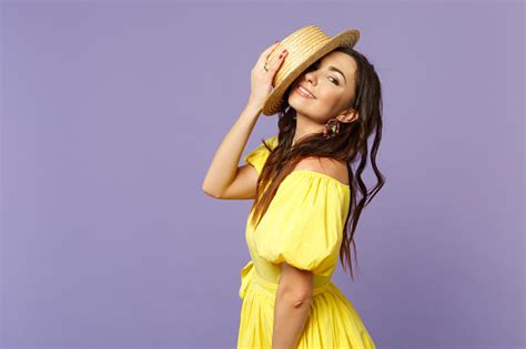 노란 드레스에 웃는 젊은 여자의 측면도 머리에 여름 모자를 들고 파스텔 바이올렛 벽 배경에 고립 된 카메라를 찾고 사람들은