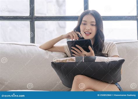 Freelance Asian Woman Surfing Browser With Tablet Sitting With Pillow On Sofa At Living Room