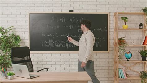 A Male Teacher Is Teaching A Chemistry Class And Explaining A Task Written On The Blackboard In