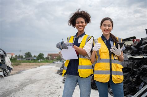 자동차 폐차장에 서서 검사하는 기계 여성 소유자 중소기업 자동차 폐차장에서 예비 부품을 선택하는 더러운 남성 수리공 스크랩 야드