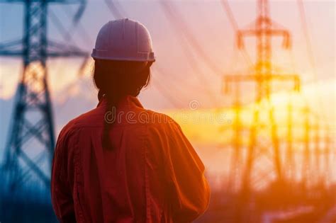 Female Engineer In Hard Hat Standing Before Power Lines At Sunset