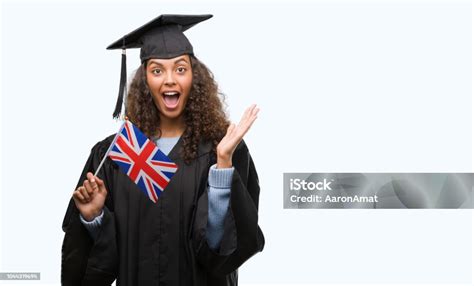 졸업 유니폼을 입고 젊은 히스패닉 여성이 큰 웃음 소리는 승리를 축 하 하는 영국 매우 행복 하 고 흥분 우승자 표현의 국기를 들고와 손을 곱슬 머리에 대한 스톡 사진 및