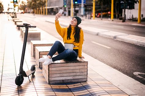 옆에 주차 된 전기 스쿠터와 대도시 지역 거리 보도에 벤치에 앉아 있는 동안 휴대 전화와 Selfie를 복용 하는 현대 여자 길거리 스타일에 대한 스톡 사진 및 기타 이미지