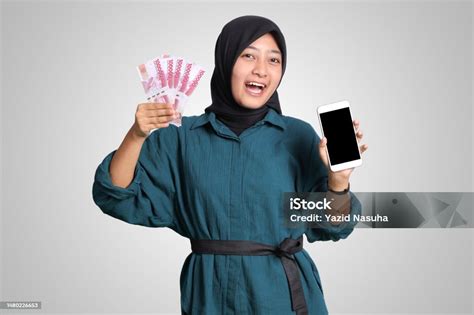 히잡을 쓴 흥분한 아시아 무슬림 여성의 초상화는 빈 화면 모형 휴대폰을 보여주면서 10만 루피아를 보여준다 재정 및 저축 개념