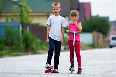 귀여운 젊은 금발의 아이들 형제 자매 스쿠터에 분홍색 옷을 입은 소녀와 햇볕이 잘 드는 거리에서 함께 노는 스케이트 보드에 잘 생긴 소년 어린이 게임과 재미있는 개념 교통