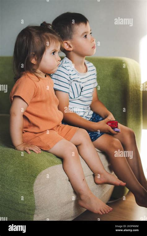 Excited Small Brother And Sister Sitting On Sofa Watching Tv In Living