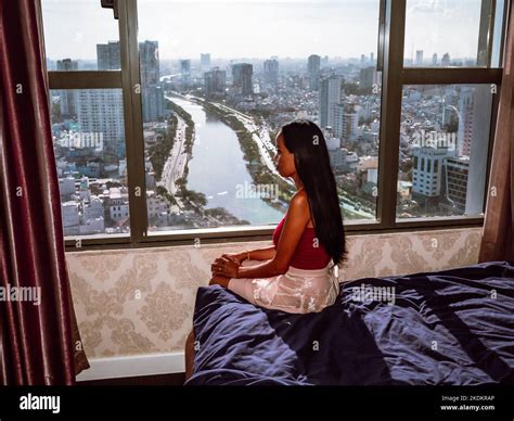 Side View Of An Asian Woman Sitting On A Bed At The Window With The View Of Ho Chi Minh Vietnam