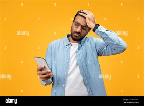A Perplexed Man In A Blue Shirt Holds His Smartphone And Looks At It