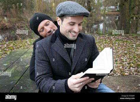 Male Gay Husbands Sit On Park Bench To Read To Each Other From Book