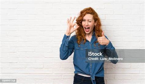 Wanita Berambut Merah Di Atas Dinding Bata Putih Menunjukkan Tanda Ok Dan Gerakan Jempol Foto