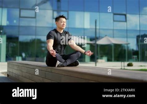 Young Adult Asian Man Meditating In The Lotus Position While Sitting On A Bench In Front Of A