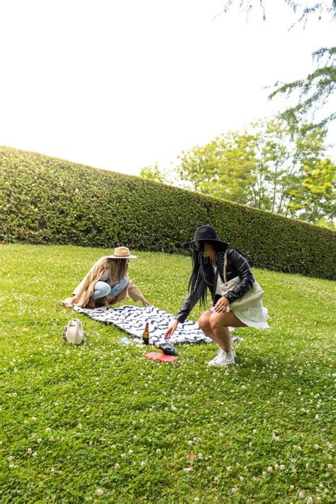 Two Friends Spreading A Blanket On The Lawn Of A Park To Spend The Day