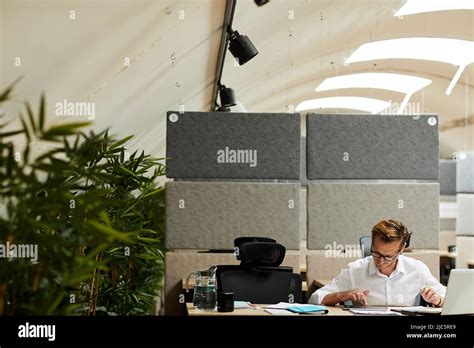 Busy Young Man In Eyeglasses Sitting At Table In Modern Office With Gradient Partitions And