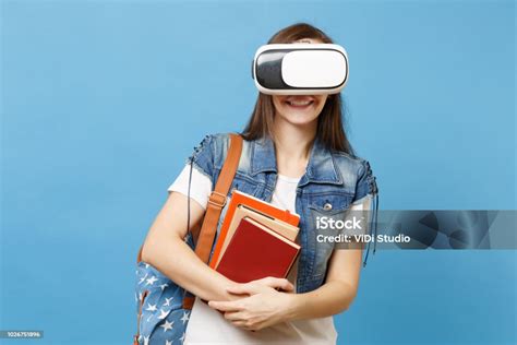 젊은 미소 여자 학생의 배낭에 가상 현실 안경 착용 데님 옷에 파란색 배경에 고립 된 학교도 서를 잡고 초상화 고등학교 대학에 교육입니다 20 29세에 대한 스톡 사진 및