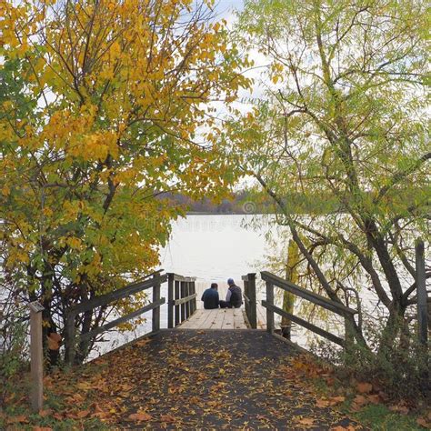 Vertical Shot Of A Couple Sitting On The Dock Surrounded By Autumn Trees Editorial Photo
